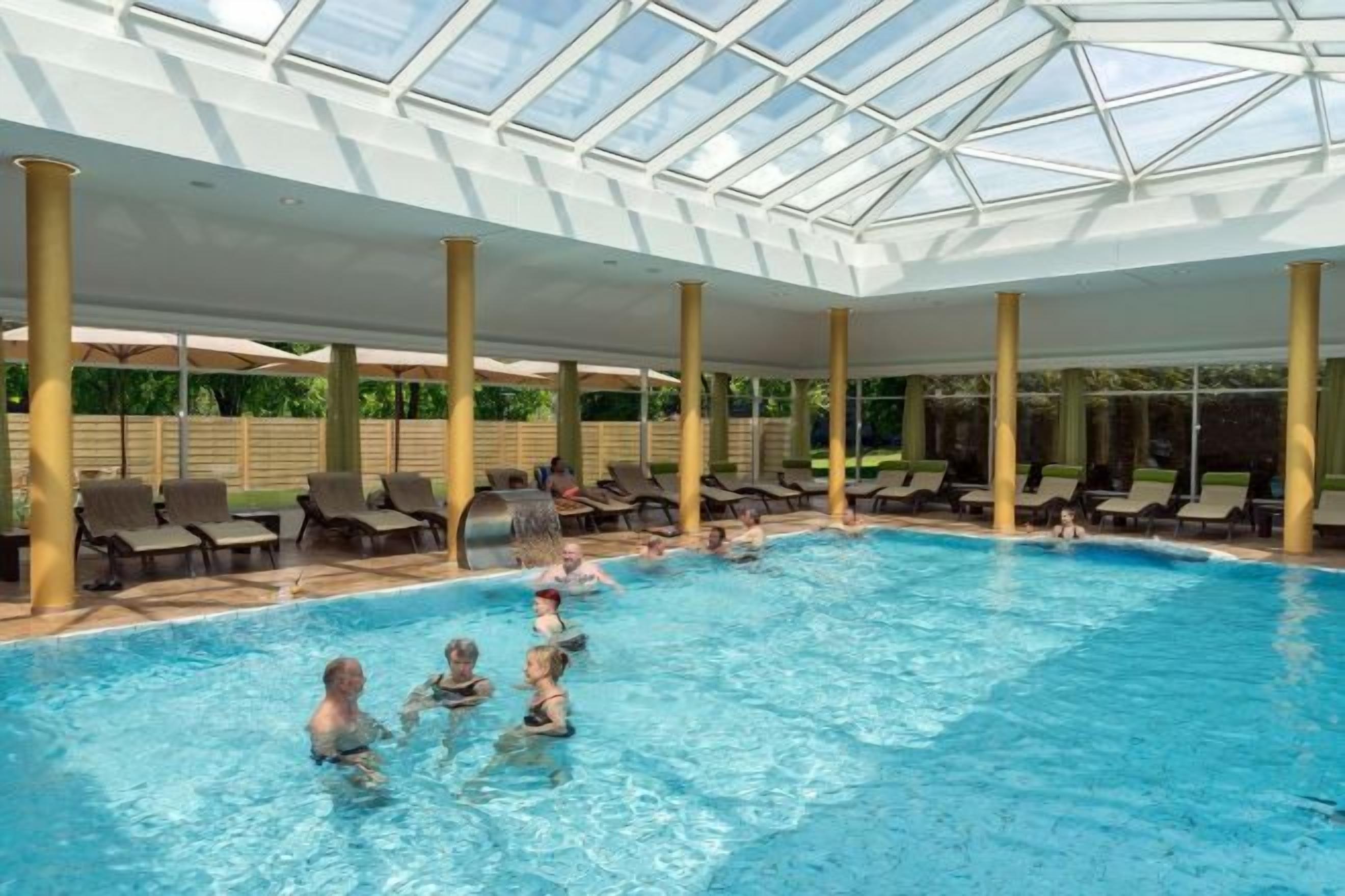 Piscina interna com teto de vidro, cadeiras de descanso e área ao redor da piscina.