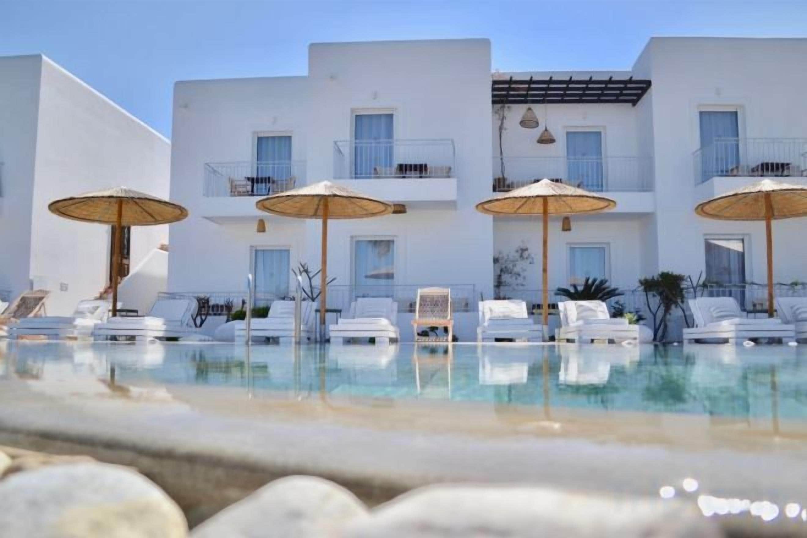 Área de piscina com espreguiçadeiras e guarda-sóis, um edifício branco com sacadas e céu claro.