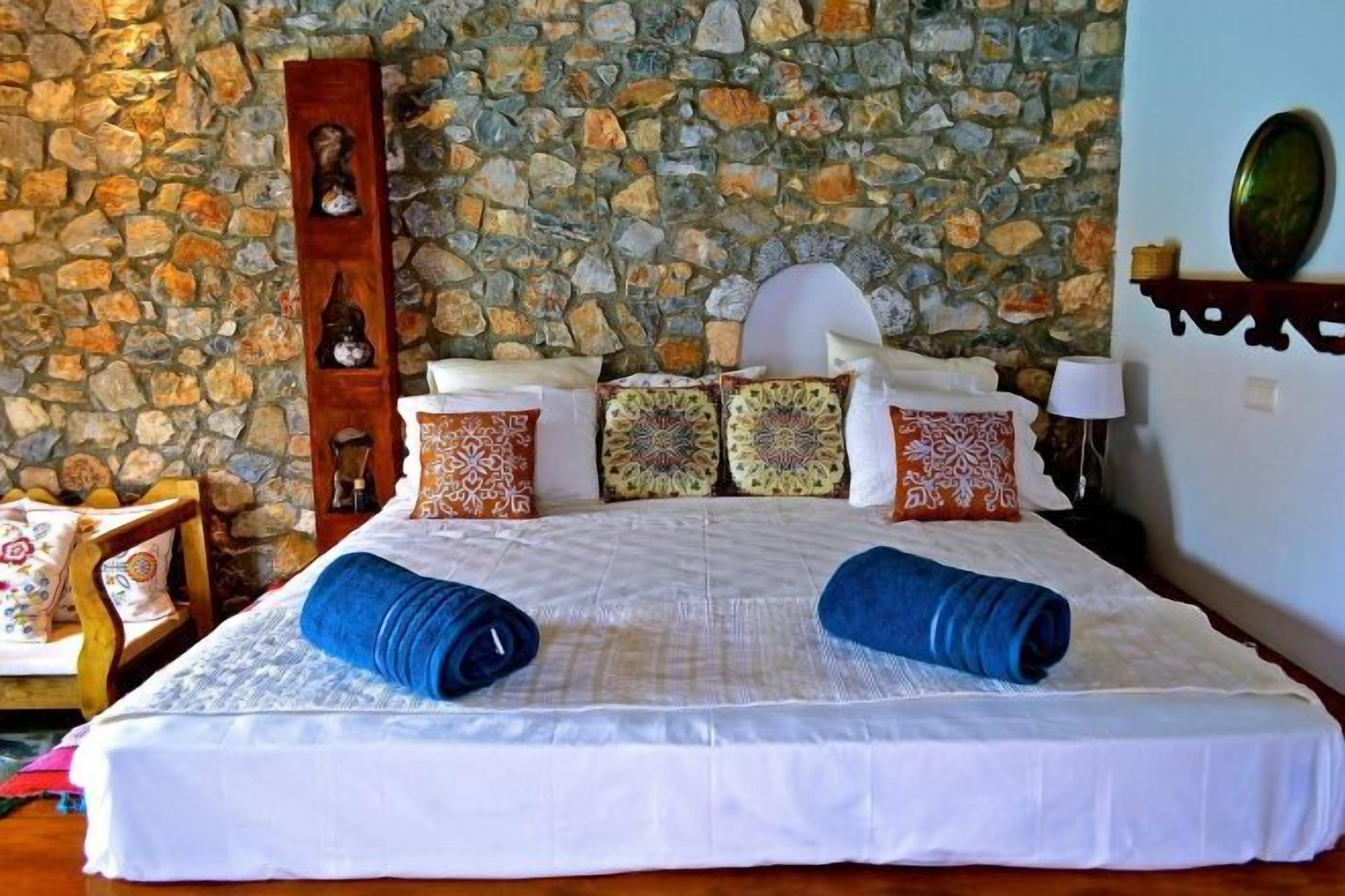 Cama com toalhas dobradas, parede de pedra e prateleira de madeira com objetos decorativos.