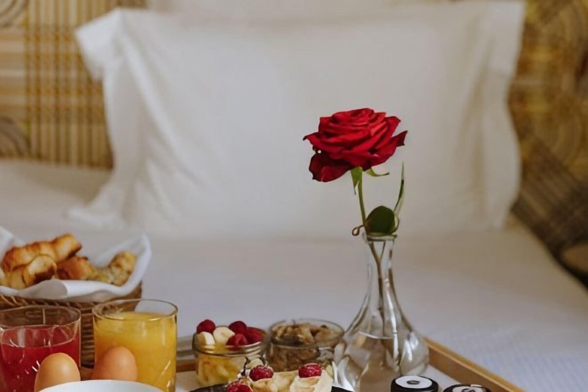 Bandeja de café da manhã com croissants, ovos e um copo de suco de laranja sobre uma cama com travesseiro branco e um vaso com uma rosa vermelha.