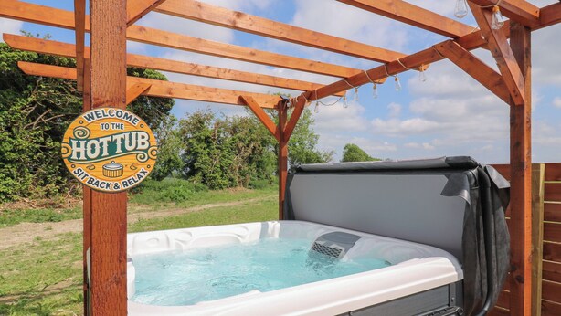 Uma banheira de hidromassagem sob uma pérgola de madeira, com uma placa que diz 'Welcome to the HOTTUB Sit Back & Relax'.