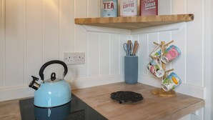 Uma cozinha com uma chaleira azul sobre um fogão por indução, uma prateleira com latas de chá, café e açúcar, e um porta-utensílios com canecas coloridas.