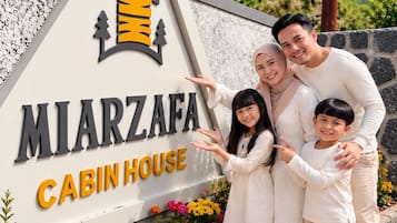 Uma família posando em frente a uma placa que diz 'Miarzafa Cabin House', com uma paisagem montanhosa ao fundo.