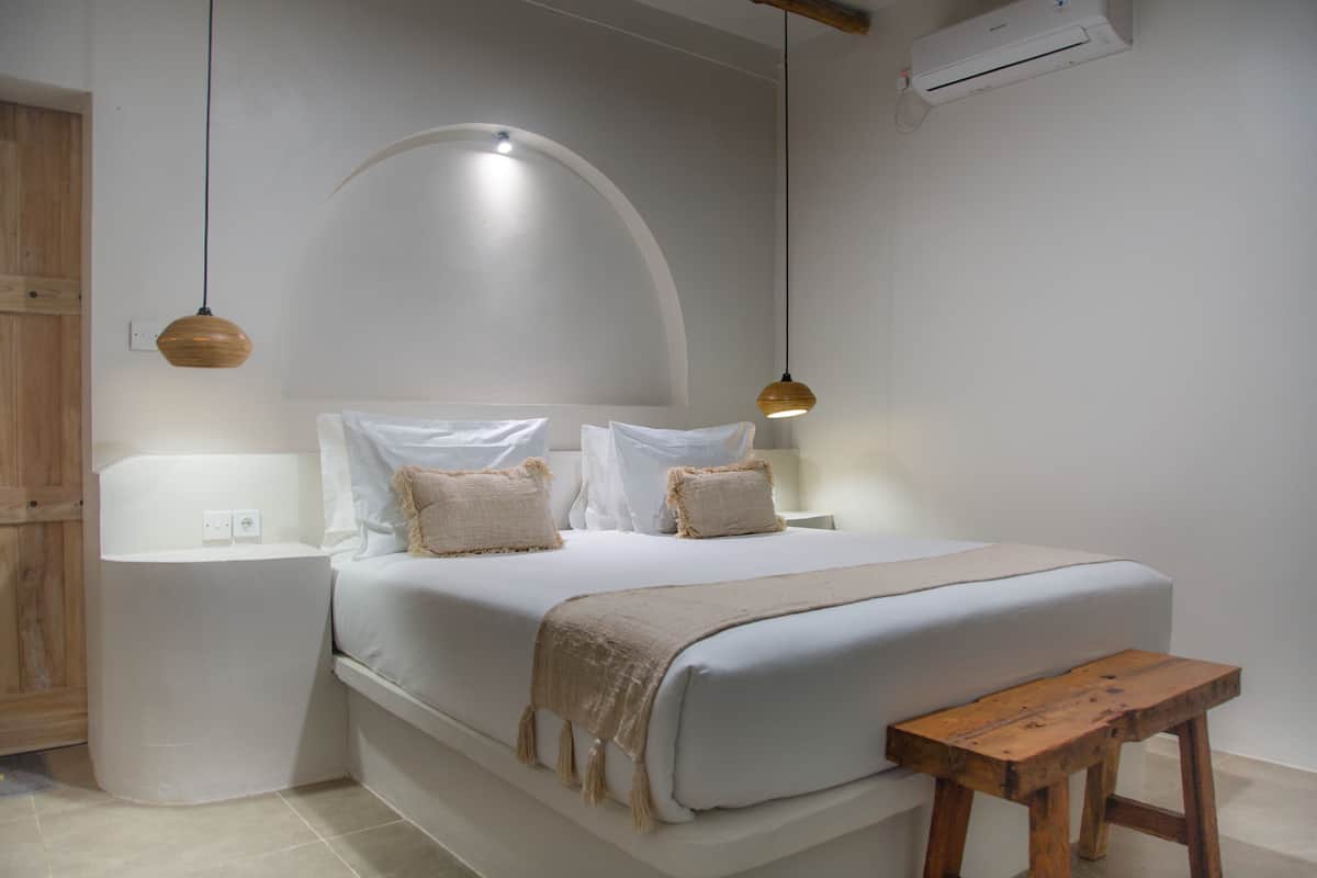 Quarto bem arrumado com cama, mesas de cabeceira, um banquinho e iluminação pendente.