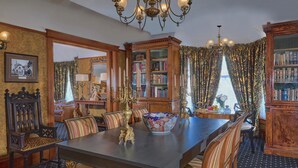 Uma sala de jantar com uma mesa comprida, cadeiras elaboradas, um candelabro e uma estante de livros.