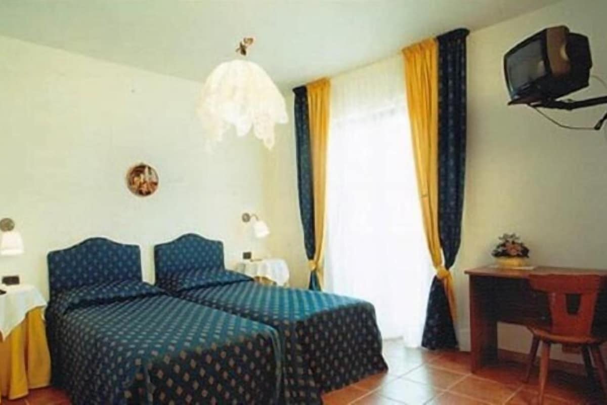 Quarto com duas camas, uma escrivaninha, televisão e janela com cortinas.