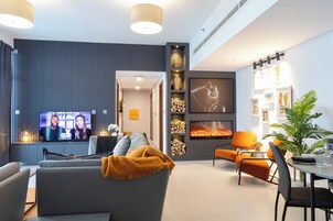 Uma sala de estar moderna com um sofá cinzento, poltronas laranja e uma mesa de jantar. Uma televisão está fixada na parede e há uma lareira com lenha à mostra.