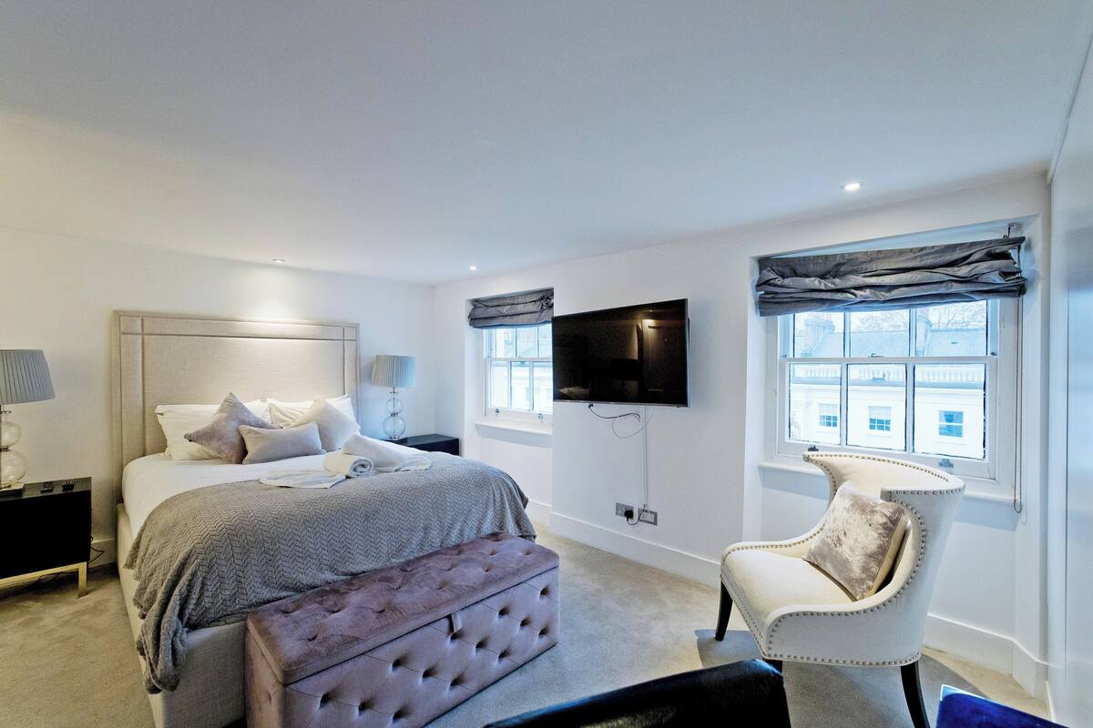Quarto moderno com uma cama grande, televisão de tela plana na parede, uma poltrona confortável e uma janela com cortinas blackout.