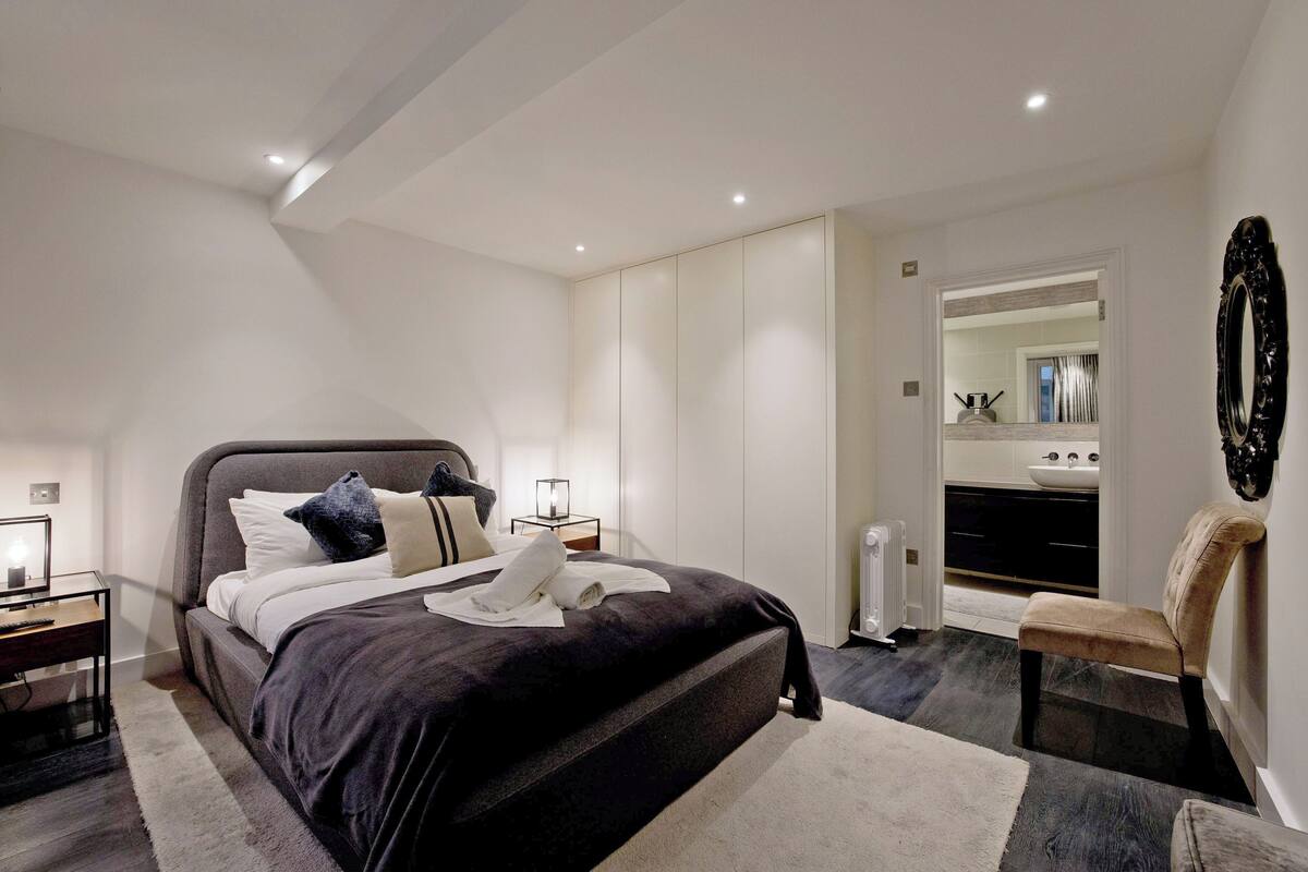 Quarto moderno com uma cama grande, mesas de cabeceira, uma cadeira e um banheiro visível através de uma porta aberta.