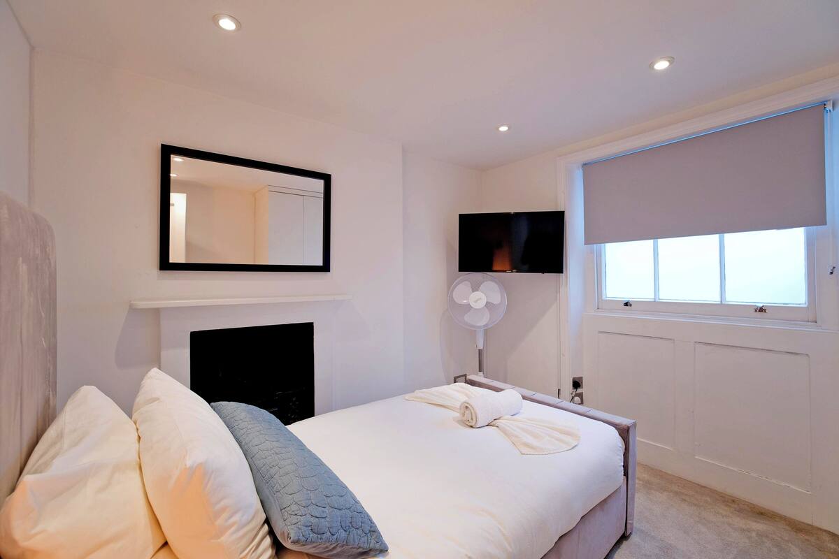 Quarto com cama, almofadas, televisão, ventilador e janela com persiana.