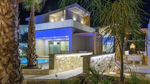 Uma casa moderna de dois andares com piscina, cercada por palmeiras e um muro de pedra.