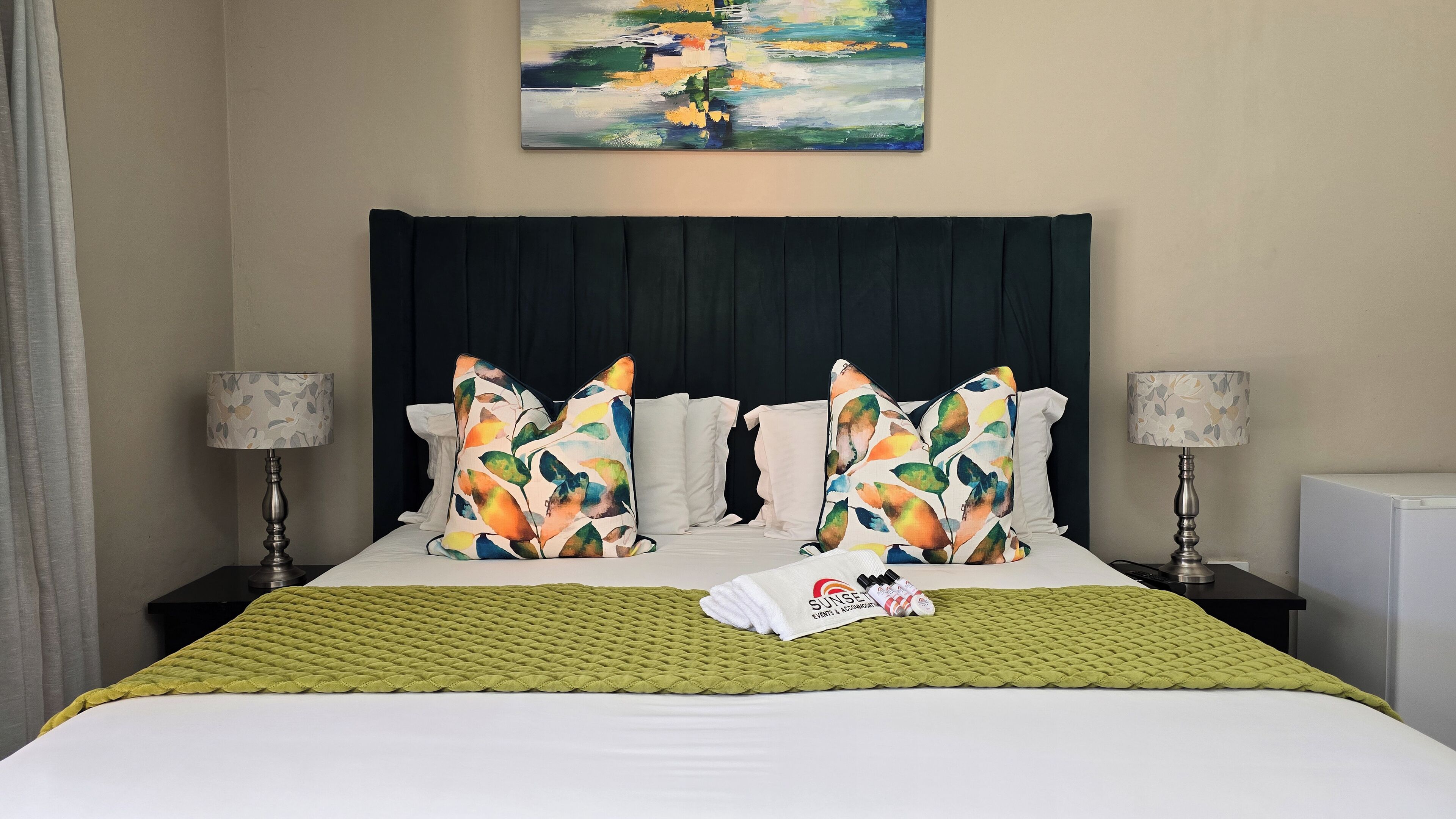 Quarto de hotel com cama, duas mesas de cabeceira, um quadro sobre a cama e um cobertor verde.