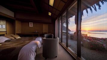 Quarto de hotel com uma janela ampla dando para um pôr do sol, uma cama, uma escrivaninha e uma cadeira.
