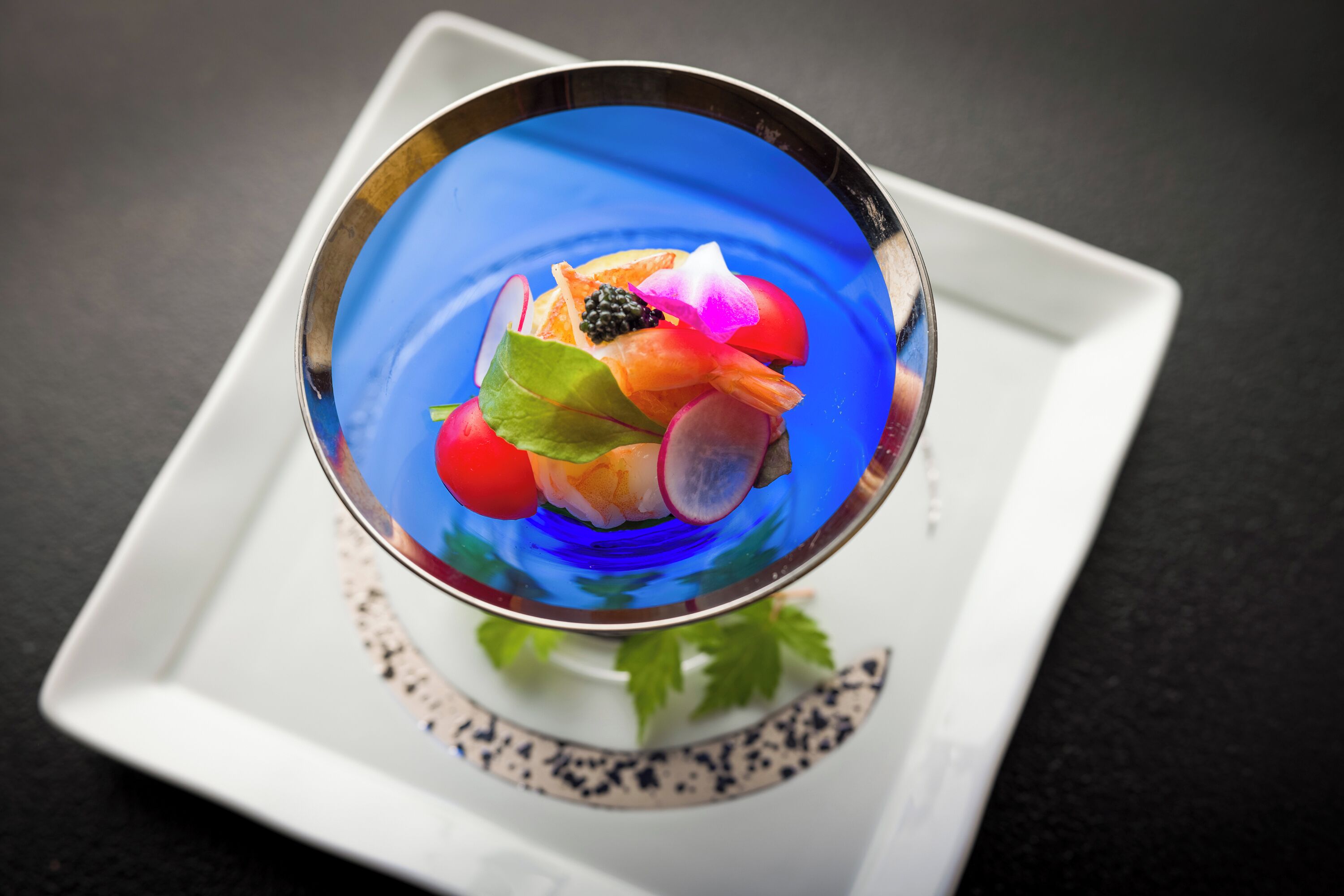 Uma travessa de vidro com borda azul sobre um prato branco, contendo um prato de frutos do mar com camarão, rabanete e um item comestível preto, possivelmente caviar, com uma folha decorativa e uma pequena fatia de laranja.