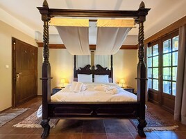 Uma cama com dossel, roupa de cama branca, um toldo, soalho de madeira e vista do exterior por grandes janelas.
