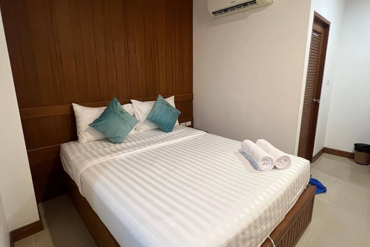 Quarto de hotel com cabeceira de madeira, cama com roupa de cama branca, duas almofadas e uma toalha enrolada sobre a cama.