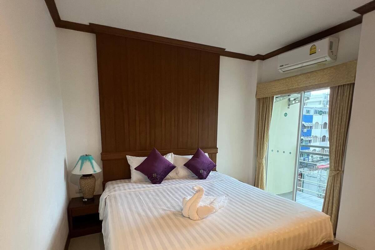 Cama bem arrumada com travesseiro e almofada em forma de cisne sobre um lençol branco, cabeceira de madeira, mesa de cabeceira com abajur e janela com cortinas.