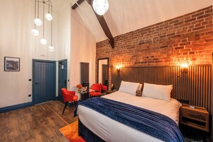 Quarto de hotel moderno com parede de tijolos, uma cama grande, uma escrivaninha com cadeira e uma cadeira vermelha.