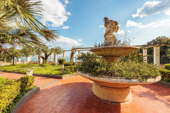 Uma fonte com uma estátua em um jardim paisagístico, com caminhos de azulejos e vegetação.