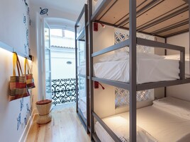Quarto com beliche, piso de madeira, uma mesa pequena com uma tigela de frutas e vista para uma varanda com guarda-corpo.