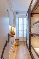 Um corredor estreito com beliches de um lado, uma porta com painel de vidro jateado e um ar condicionado fixado na parede.