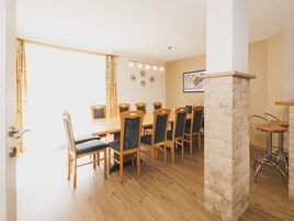 Uma área de refeições com mesa e cadeiras de madeira, um pilar de pedra e um balcão com banquetas.