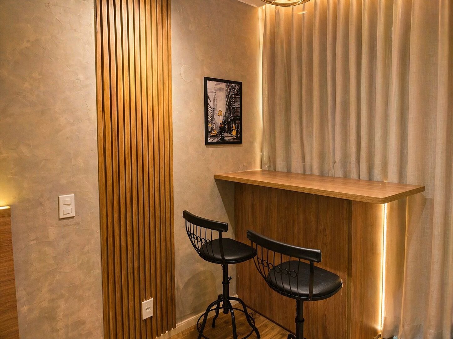 Quarto de canto com parede de ripas de madeira, um quadro emoldurado, balcão de madeira e duas cadeiras pretas.