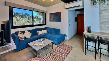 Sala moderna com sofá azul, mesa de centro e televisão. Há uma janela com vista para árvores, um quadro na parede e um espaço de refeições com cadeiras.