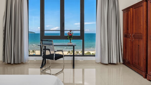 Quarto com uma grande janela com vista para uma praia, uma cadeira, uma pequena mesa com um vaso e um guarda-roupa de madeira.