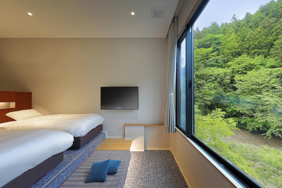 Quarto de hotel moderno com duas camas, uma televisão de tela plana e uma janela grande com vista para uma área arborizada.
