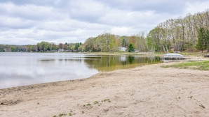 Vista para o lago com uma praia de areia, um píer e uma casa ao fundo.