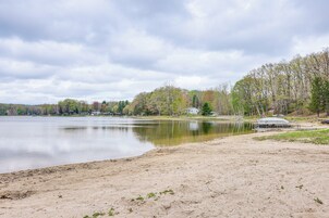 Vista para o lago com uma praia de areia, um píer e uma casa ao fundo.