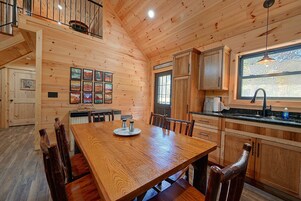 Interior de uma cabana de madeira, com mesa de jantar, cadeiras, cozinha e escada para um andar superior.