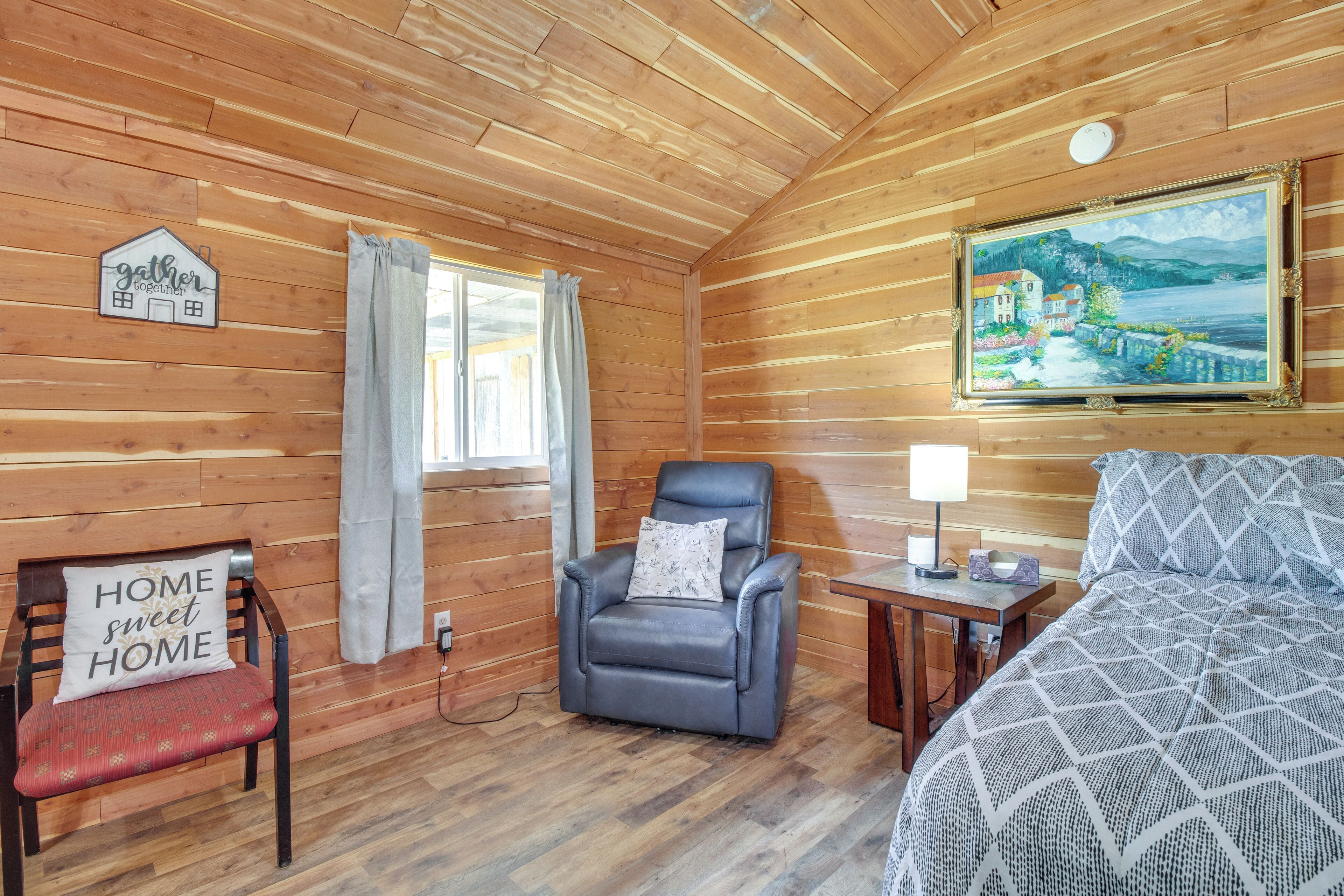 Um quarto aconchegante com paredes de madeira, uma poltrona confortável, uma cama, um quadro e uma janela com cortinas.