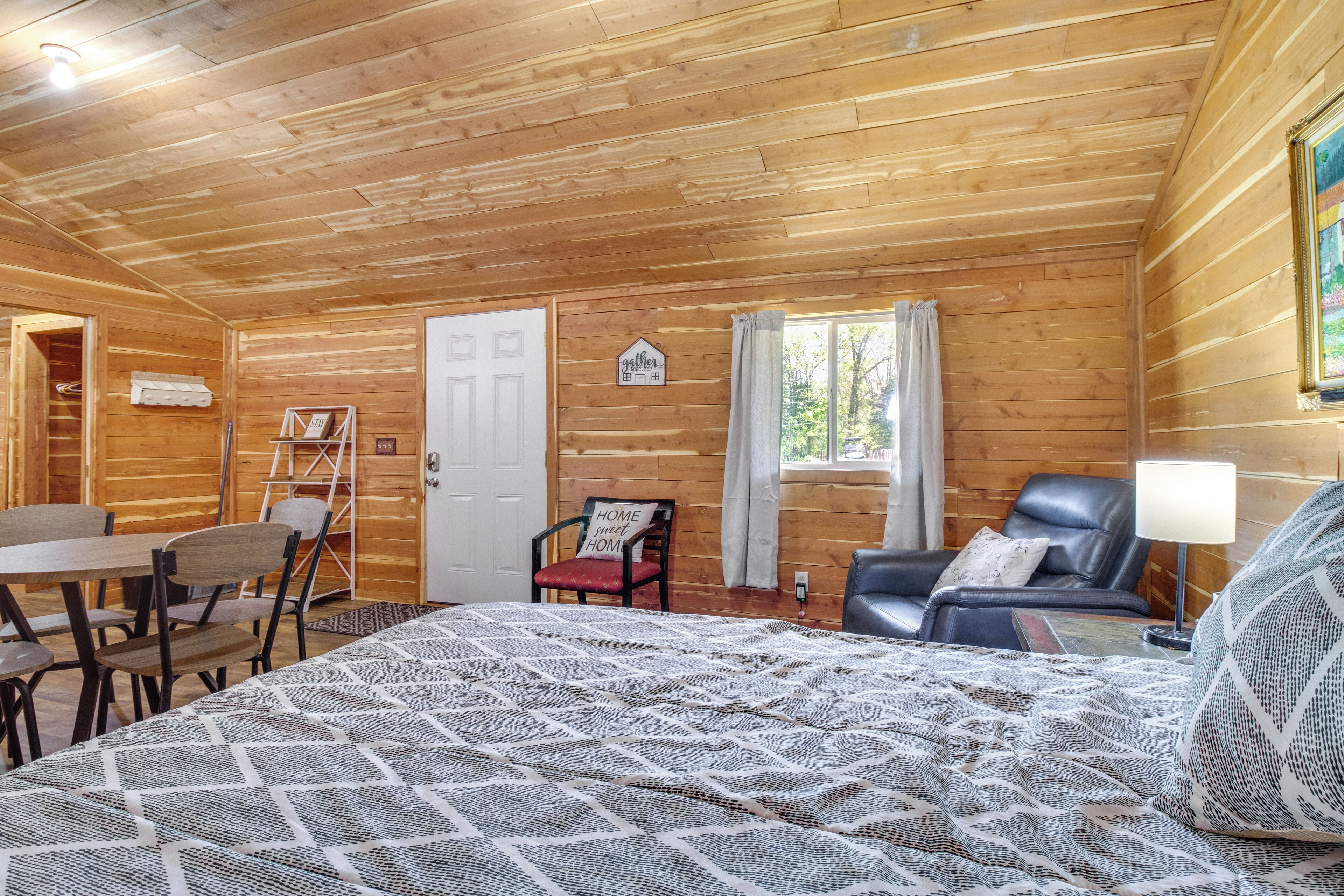 Um quarto de cabana de madeira acolhedor, com uma cama, uma mesa de jantar com cadeiras, uma poltrona com almofada e uma poltrona de couro preto. Há também uma janela com cortinas e uma porta.