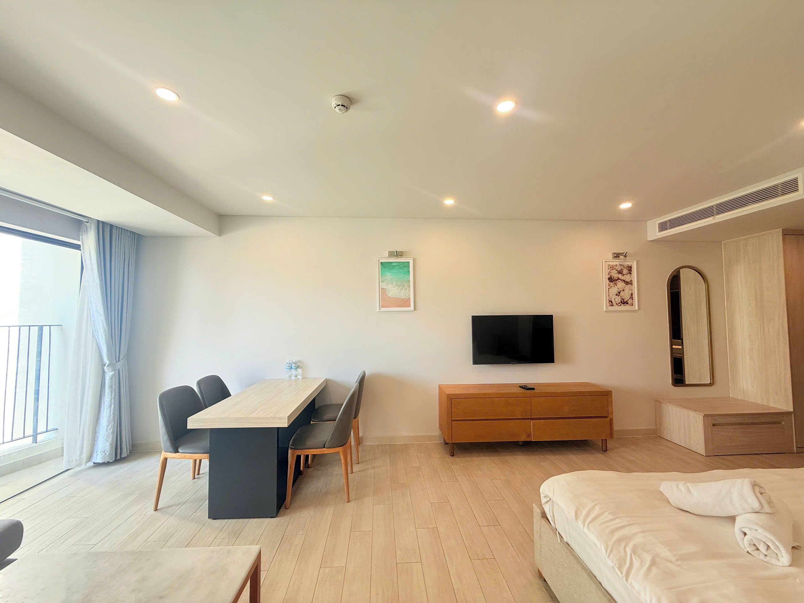 Quarto de hotel com cama, televisão, mesa de jantar com cadeiras, armário de madeira e uma janela grande com cortinas.