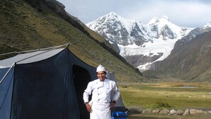 Um chef em frente a uma tenda, com uma paisagem montanhosa ao fundo.