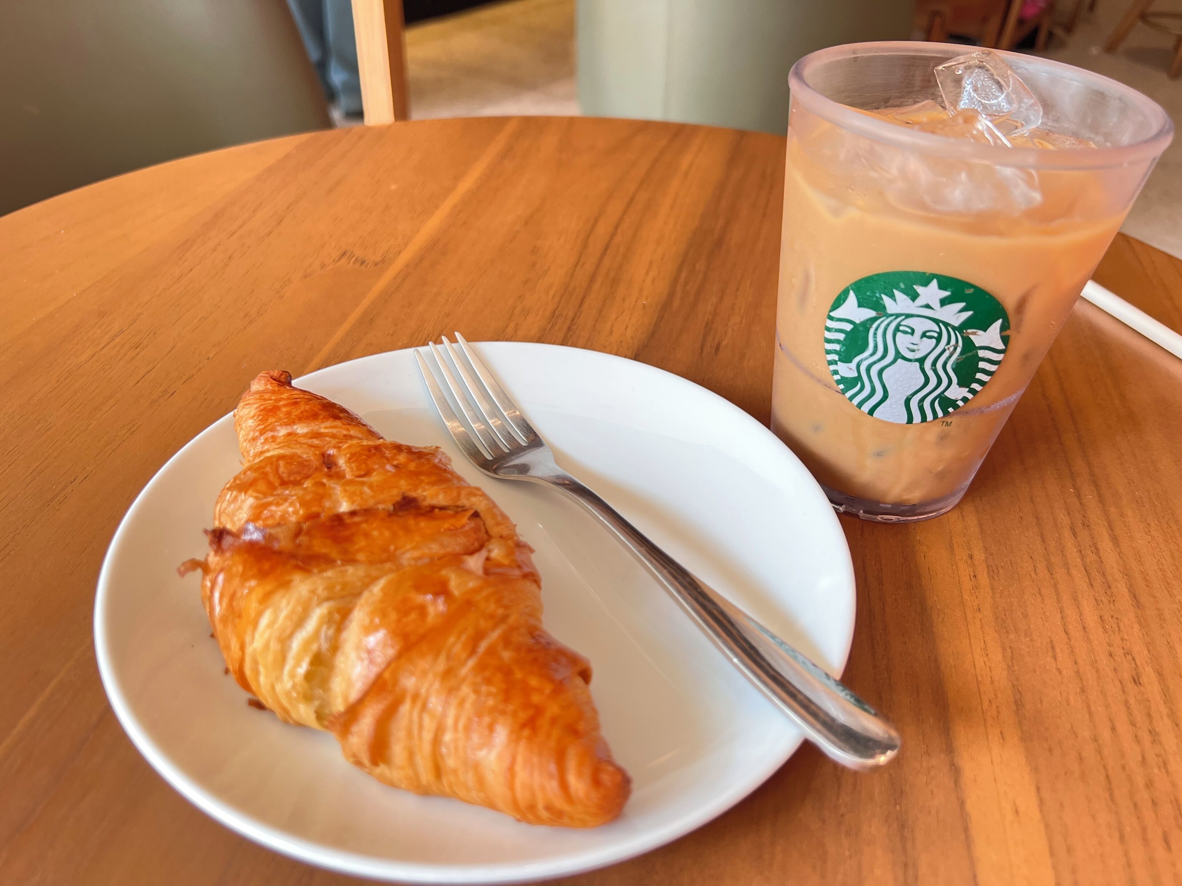 Um croissant sobre um prato branco, com um garfo ao lado de uma chávena de café gelado Starbucks.