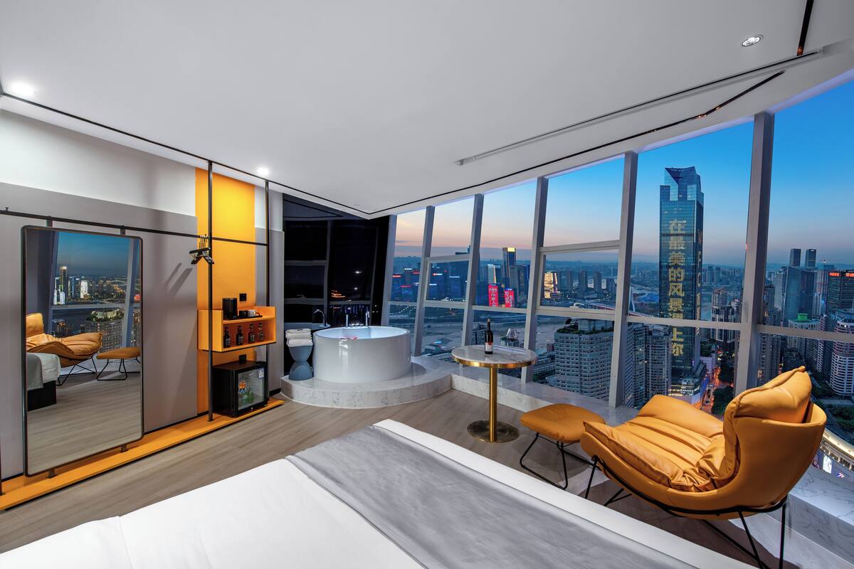 Quarto de hotel moderno com uma cama grande, área de estar com vista para a cidade, uma pequena mesa redonda e uma janela ampla que oferece uma vista panorâmica da paisagem urbana.
