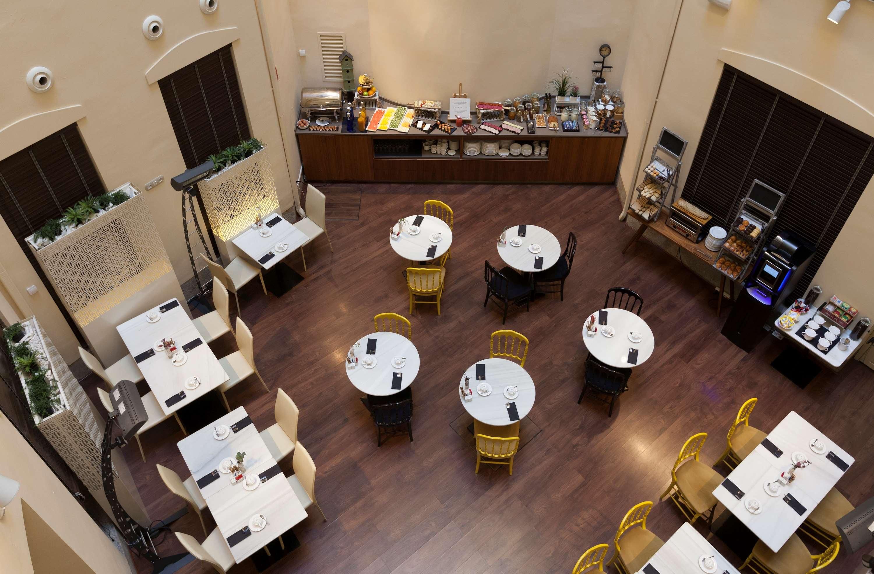 Espaço de refeições de hotel com várias mesas e cadeiras, um buffet com diversos alimentos e plantas decorativas.
