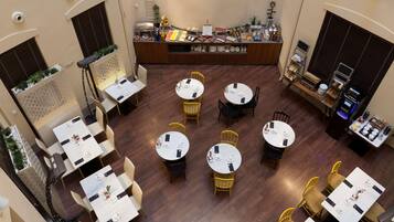 Espaço de refeições de hotel com várias mesas e cadeiras, um buffet com diversos alimentos e plantas decorativas.