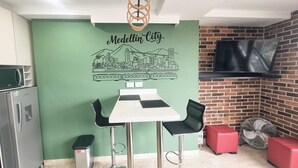 Uma cozinha compacta com micro-ondas, geladeira, mesa com duas cadeiras, lixeira, ventilador e TV fixada em uma parede de tijolos.