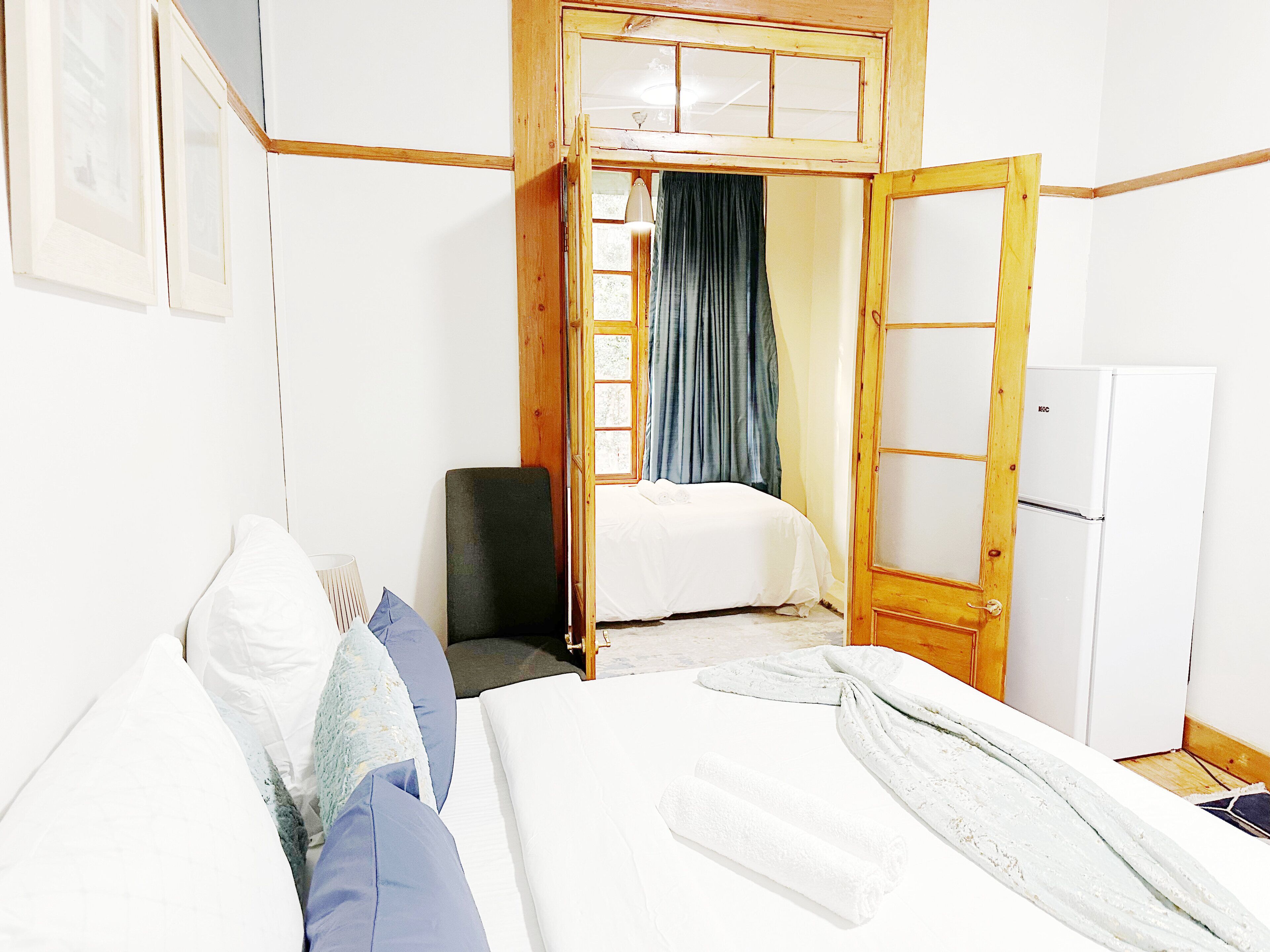 Quarto de hotel com cama, uma cadeira, escrivaninha, geladeira e janela com cortinas.