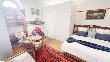 Quarto com cama, uma cadeira, uma mesinha com um vaso, um tapete e um quadro na parede.
