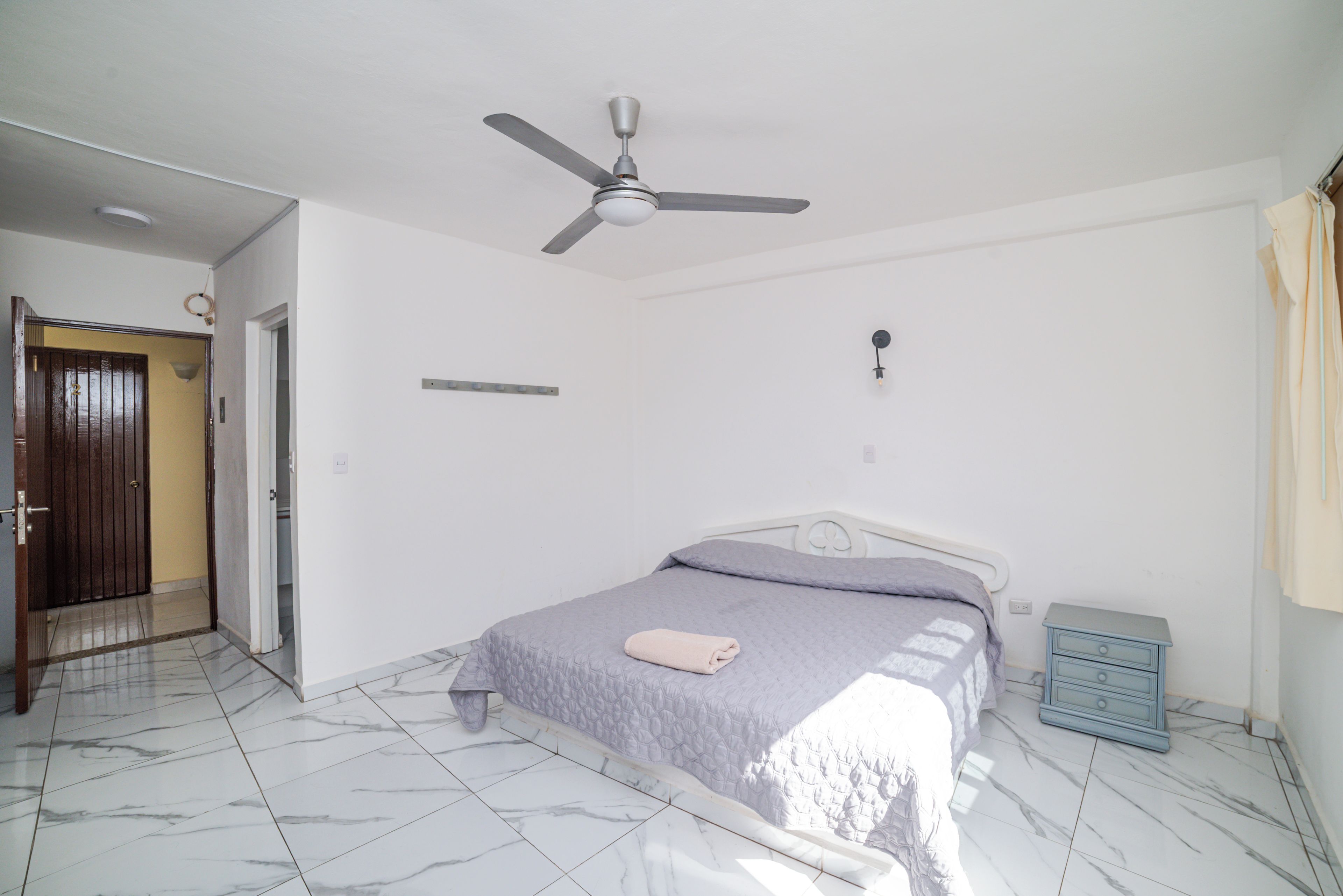 Quarto com cama, ventilador de teto, mesa de cabeceira, porta e interruptor de luz na parede.