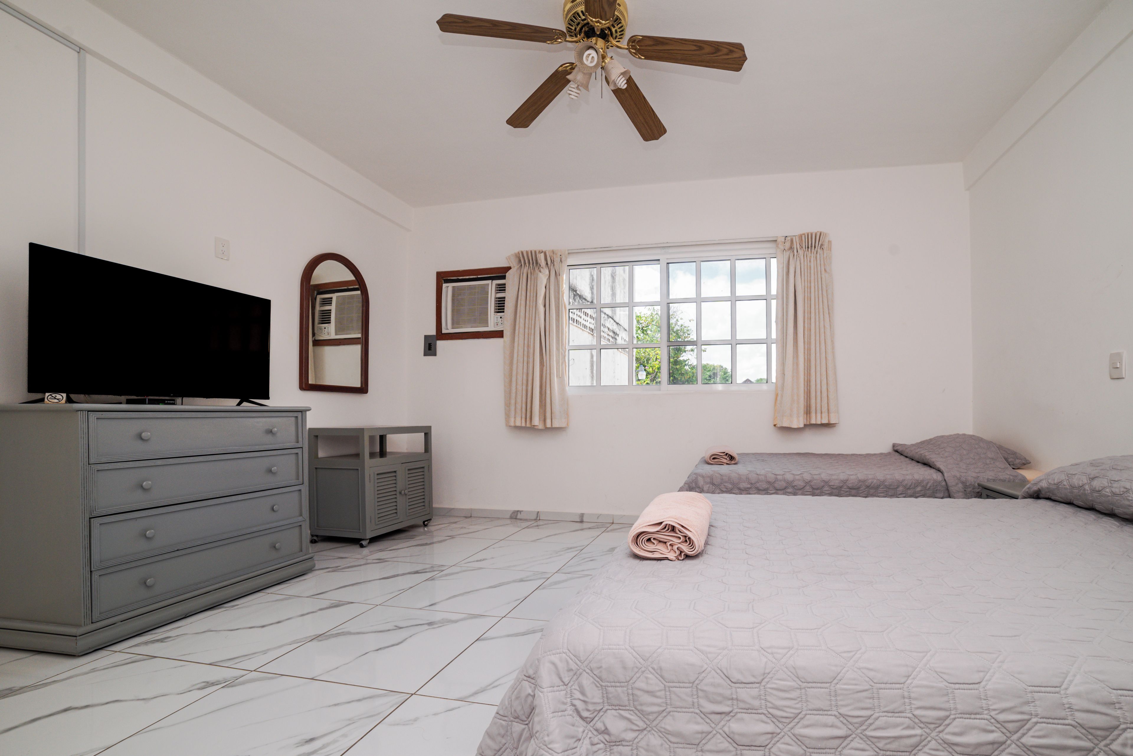 Quarto com cama, televisão, penteadeira, ventilador de teto e janela com cortinas.