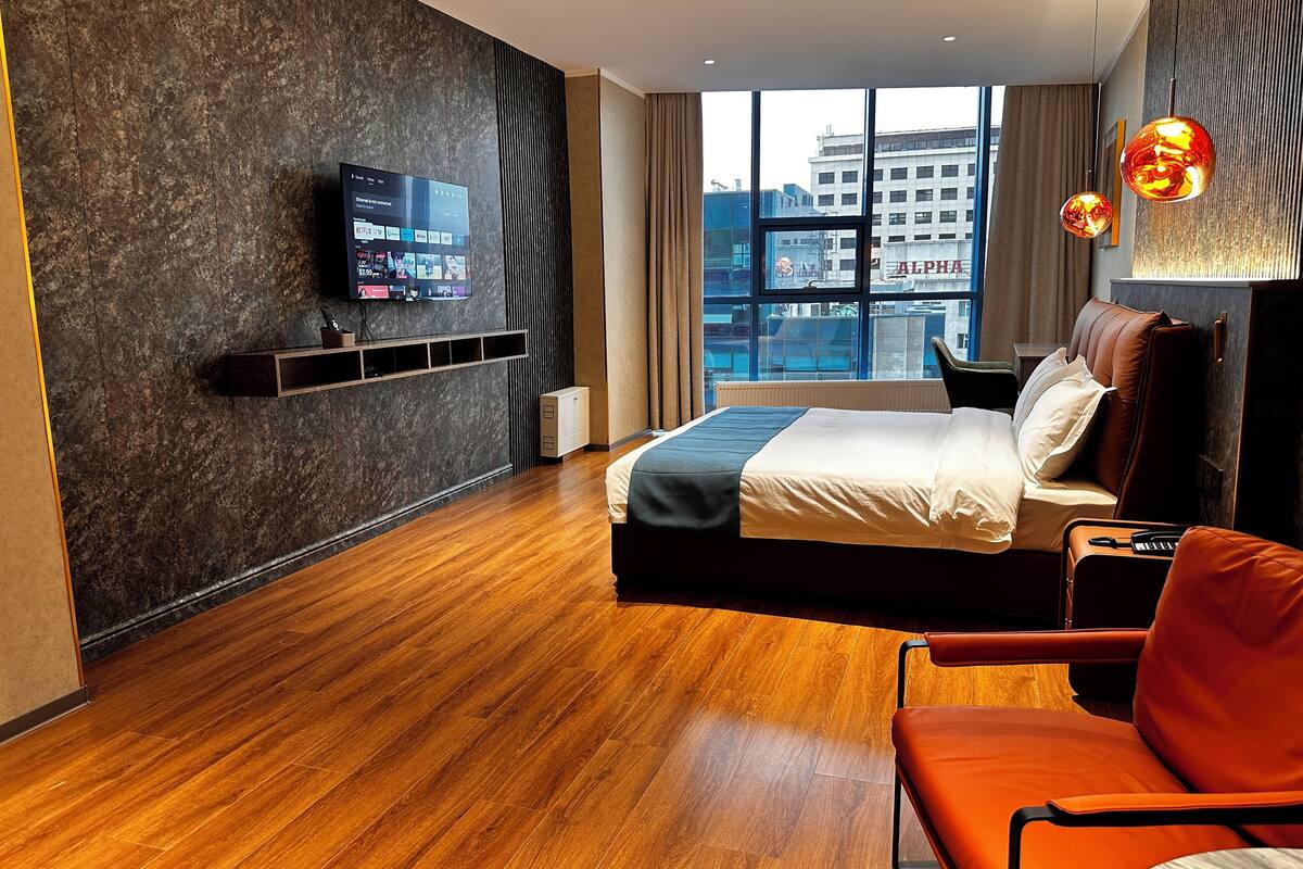 Quarto de hotel moderno com uma cama grande, TV fixada na parede e vista da cidade através de amplas janelas.