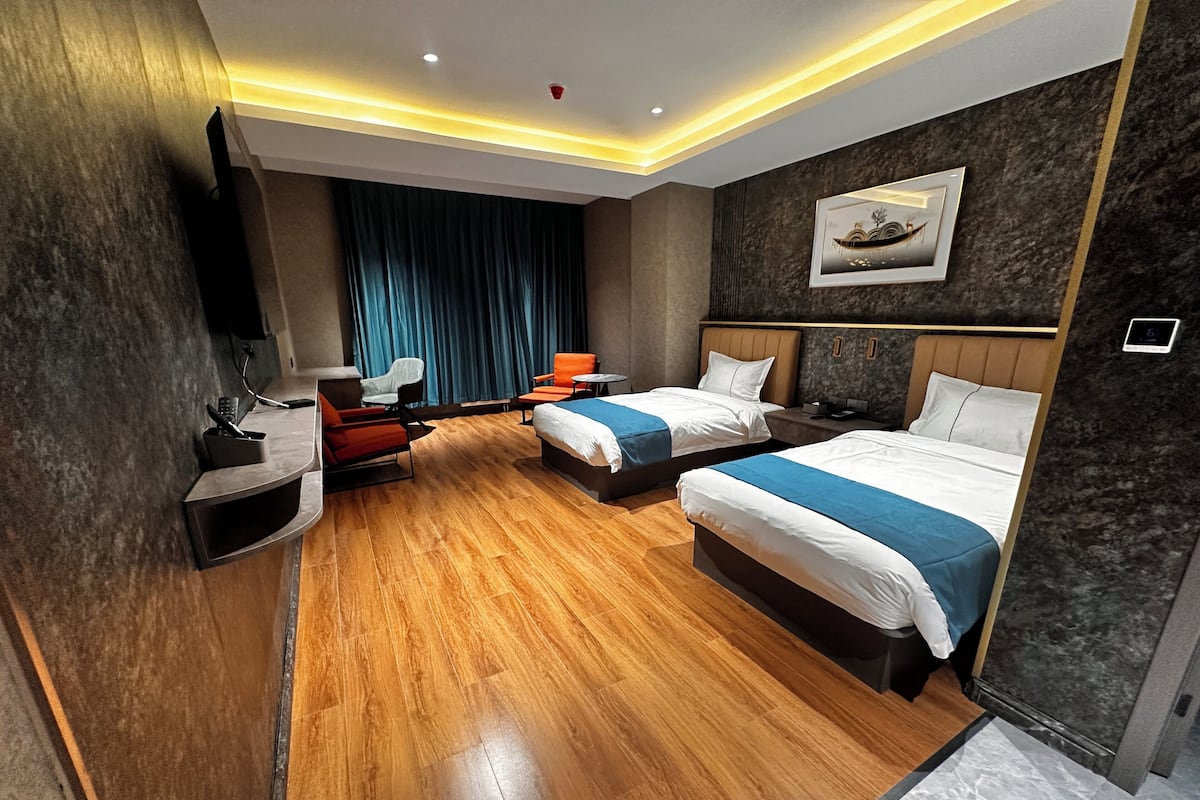 Quarto de hotel moderno com duas camas, uma escrivaninha, uma cadeira, televisão e uma janela grande com cortinas.