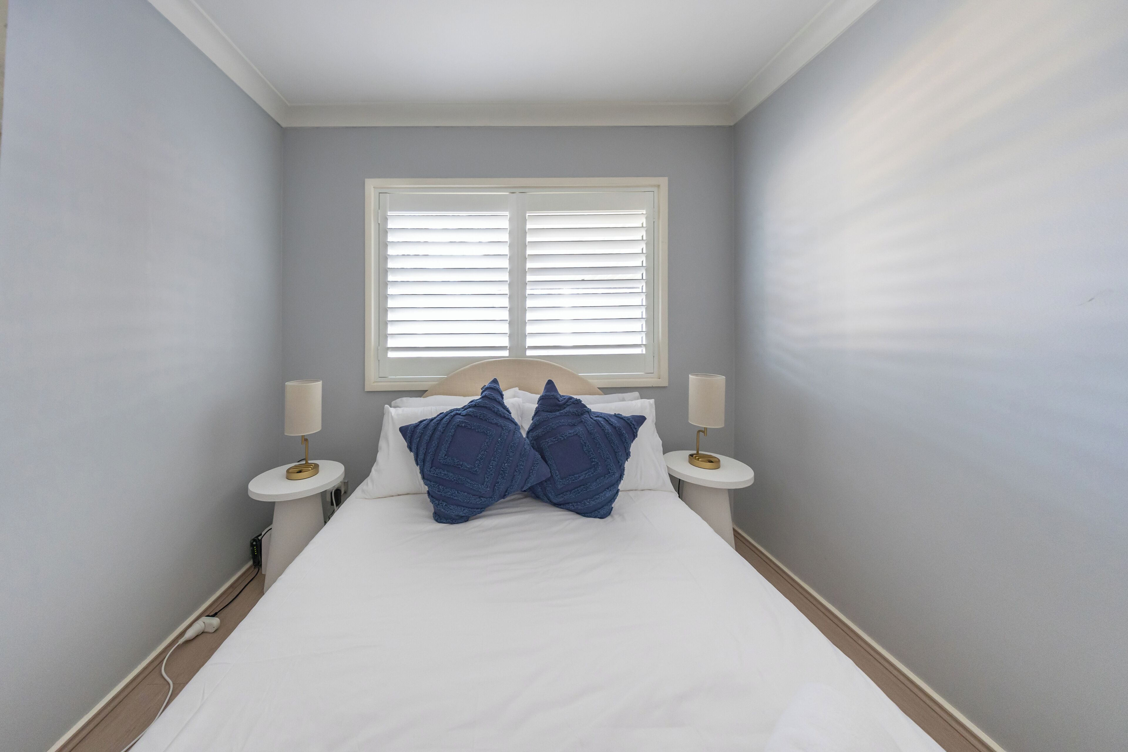 Cama bem arrumada com roupa de cama branca e duas almofadas azuis decorativas, com mesas de cabeceira com abajures de cada lado, em um quarto com paredes cinza claro e uma janela com persianas brancas.