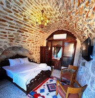Quarto com parede de pedra, cama com cabeceira de madeira, uma cadeira de madeira, um espelho e uma televisão.
